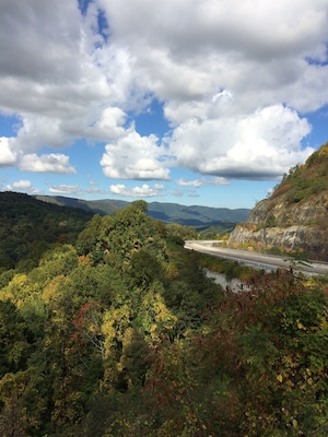 Uma estrada sinuosa serpenteia por um vale arborizado, com um rio e montanhas ao longe, sob um céu parcialmente nublado.