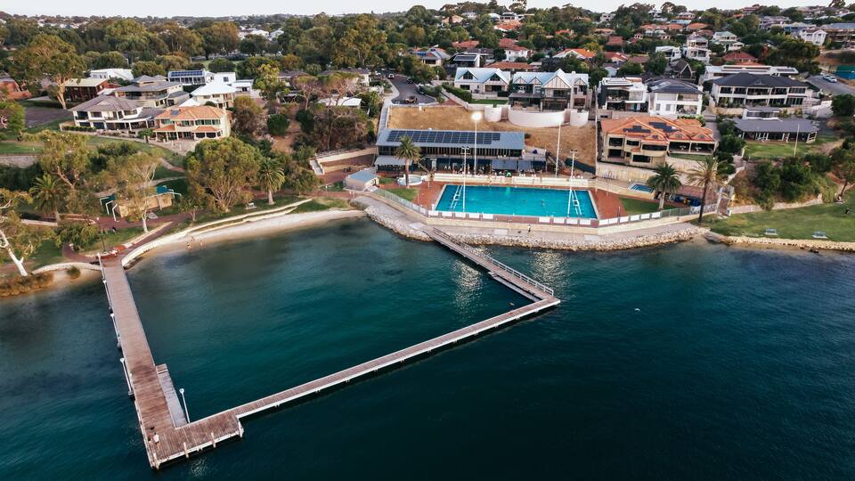 Vista aérea de uma área residencial à beira-mar, com uma grande piscina, um píer de madeira e casas equipadas com painéis solares.