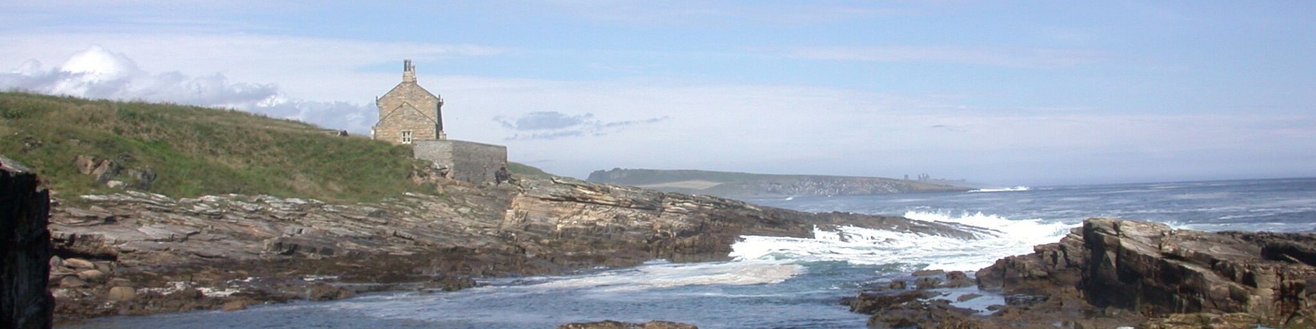Paisagem costeira com falésias rochosas, ondas quebrando nas rochas e um edifício de pedra numa colina gramada.