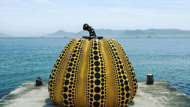 Uma escultura grande, amarela e preta com bolinhas, sobre um platô de concreto à beira-mar.