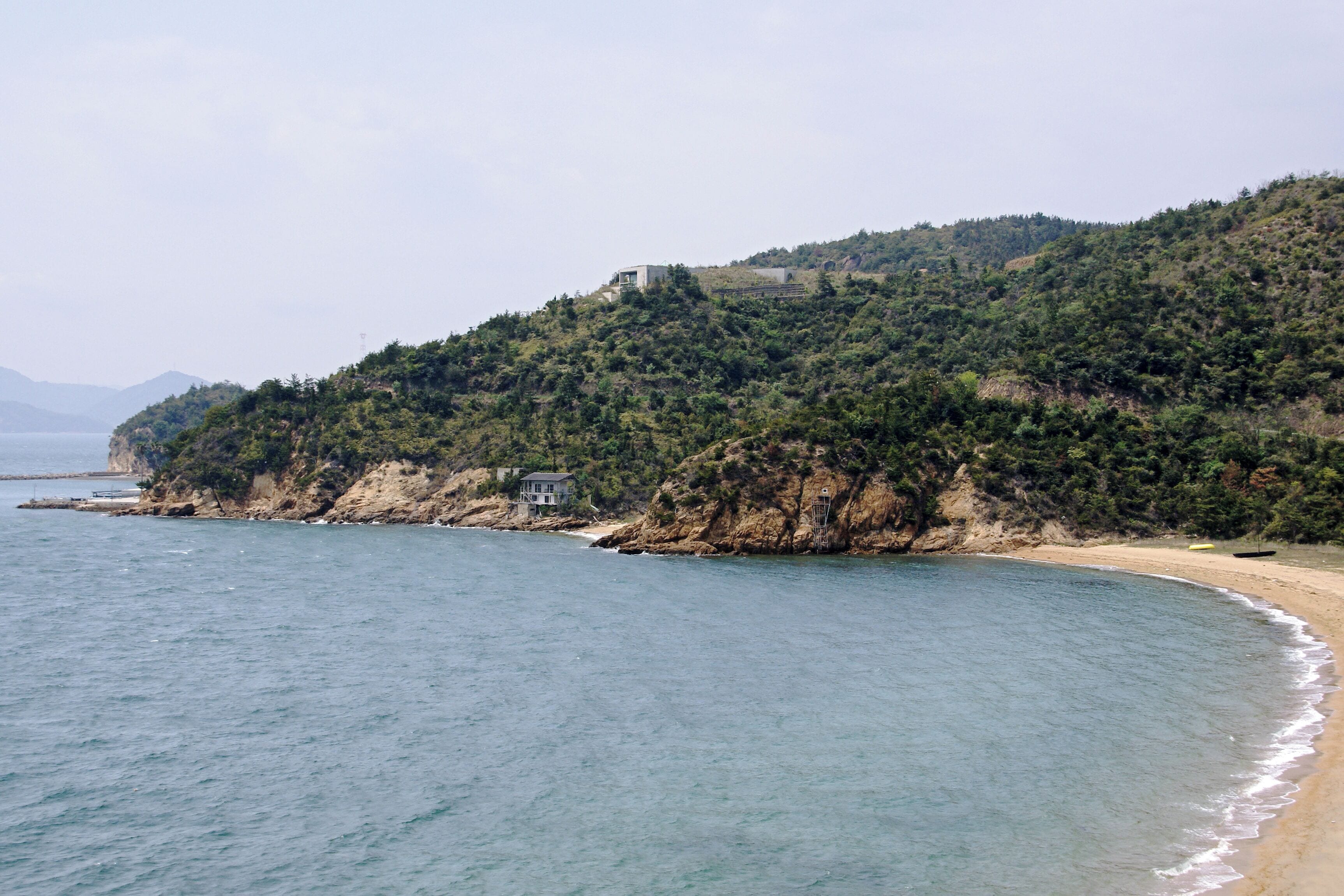 Paisagem costeira com praia, falésia coberta de vegetação e um edifício no topo da colina.