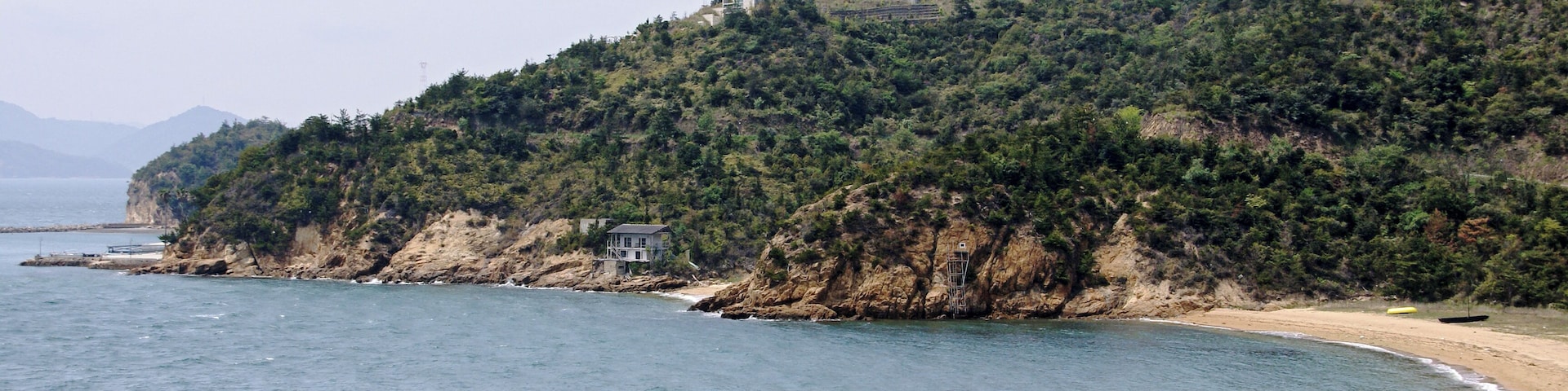 Paisagem costeira com praia, falésia coberta de vegetação e um edifício no topo da colina.
