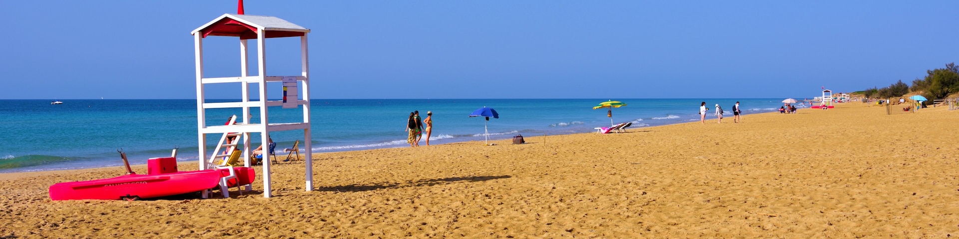 Uma praia arenosa com uma torre salva-vidas, posto de salva-vidas vermelho e algumas pessoas à distância.
