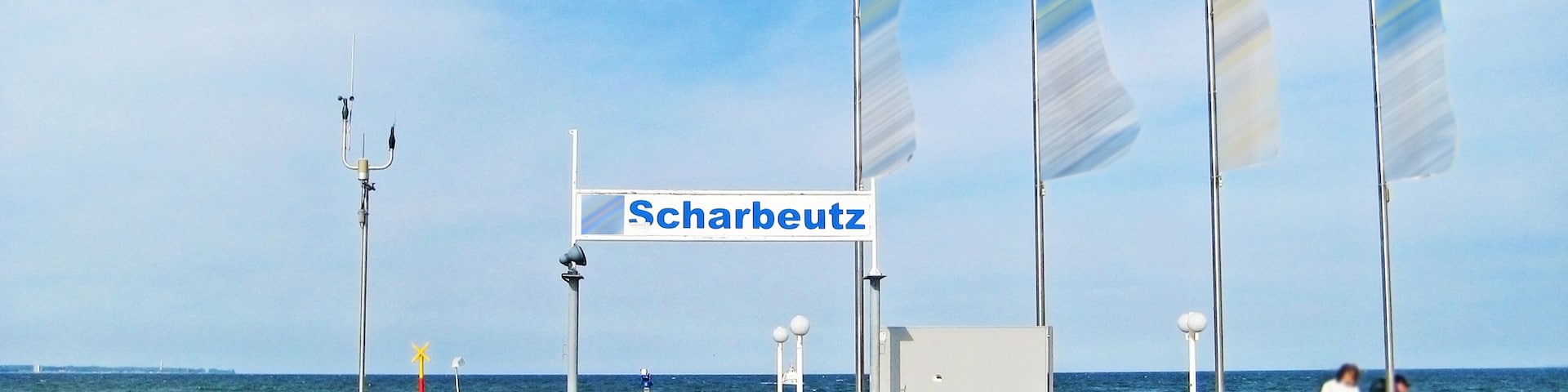 Um cais de madeira que se estende sobre a água, com uma placa indicando 'Scharbeutz' e bandeiras em mastros.
