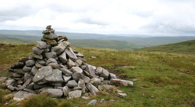 Um cairn numa colina gramada com vista panorâmica de colinas ondulantes ao fundo.