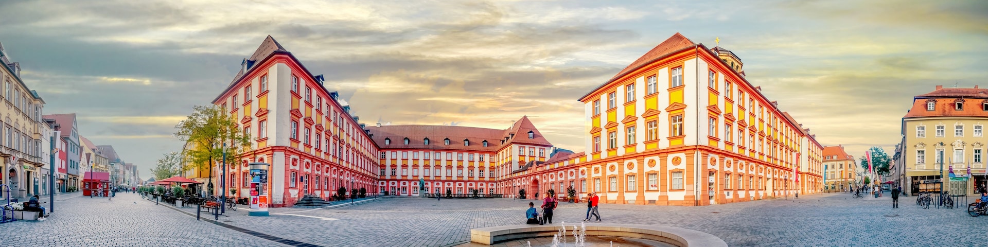 Uma praça histórica com calçamento de pedra, uma fonte central e edifícios coloridos com arquitetura tradicional.