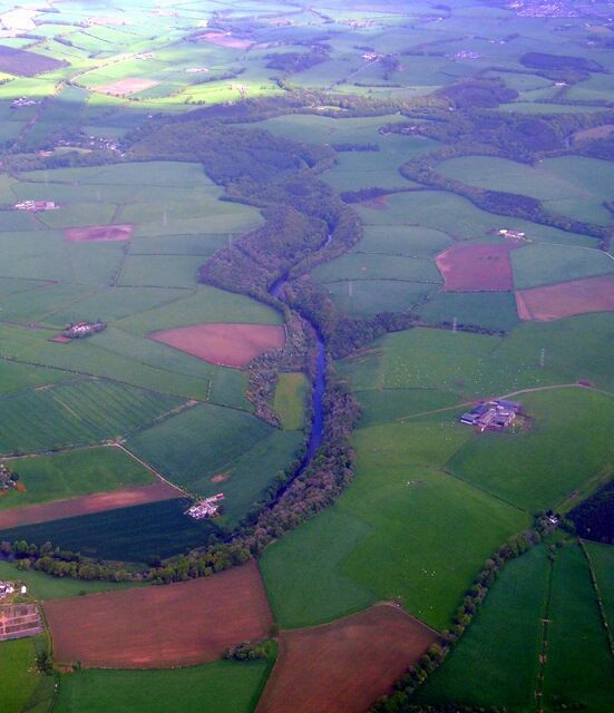 Vista aérea de um rio serpenteando por campos agrícolas, com áreas de floresta e pequenos edifícios.