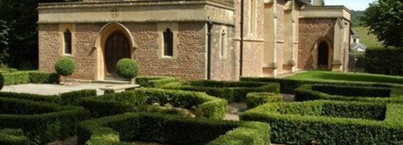 Uma igreja com um campanário, rodeada por um jardim formal com sebes e um caminho de cascalho.