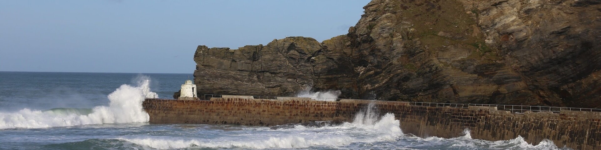 Um litoral rochoso com ondas quebrando contra um muro marítimo e um farol no topo de um penhasco.