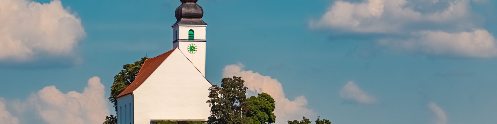 Uma igreja branca com uma cruz verde na torre, rodeada por árvores e casas.