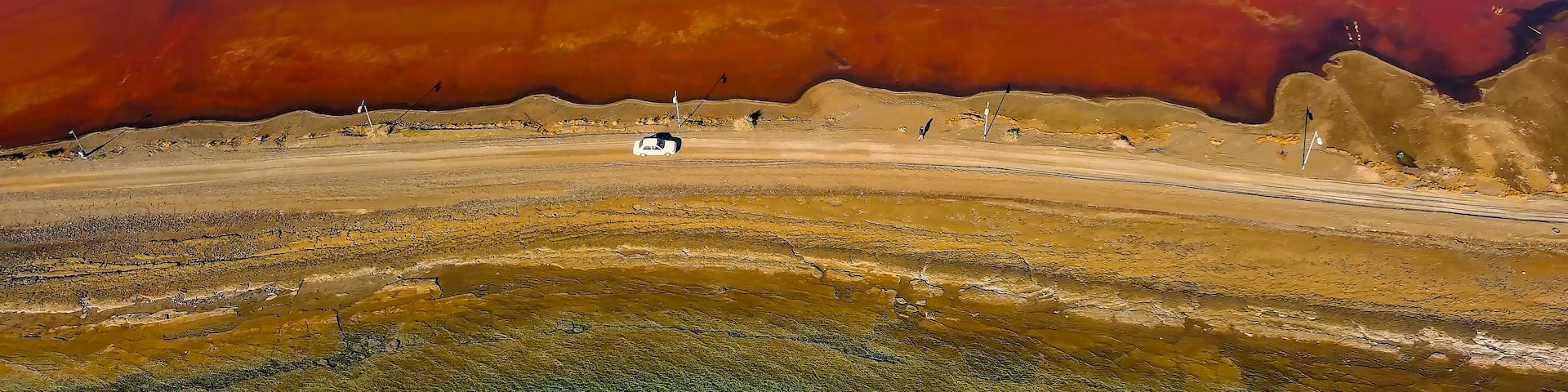 Paisagem desértica com veículo branco, corpos d'água límpidos e céu acastanhado avermelhado.