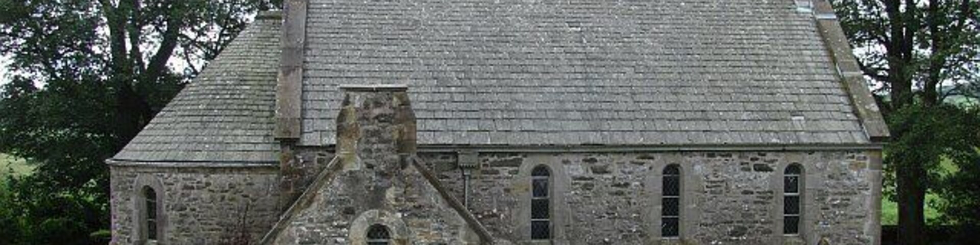 Uma igreja de pedra com um telhado íngreme e um campanário, rodeada por um espaço gramado e árvores.