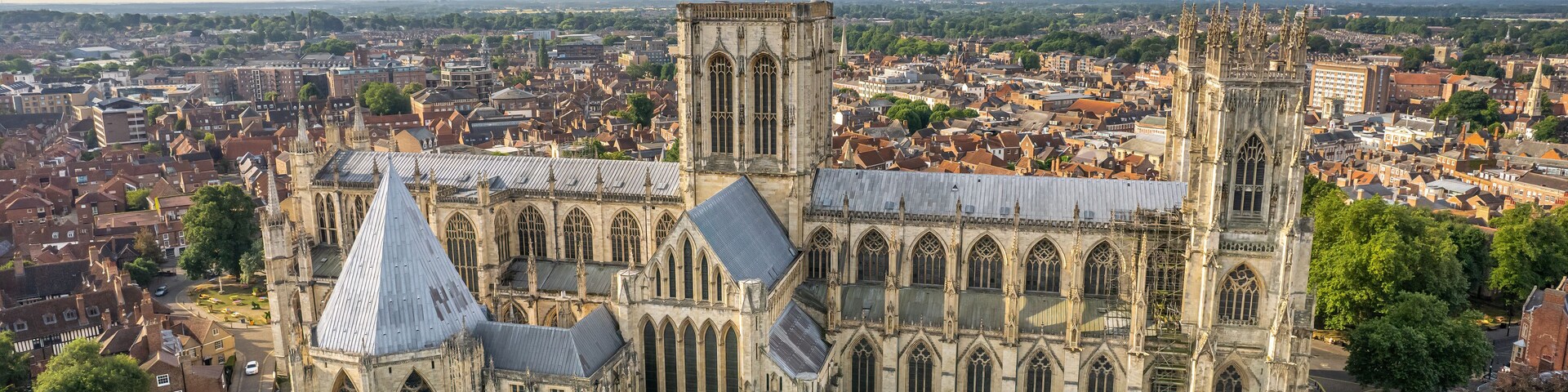Uma grande catedral gótica com uma torre imponente, rodeada por um cenário urbano e vegetação.