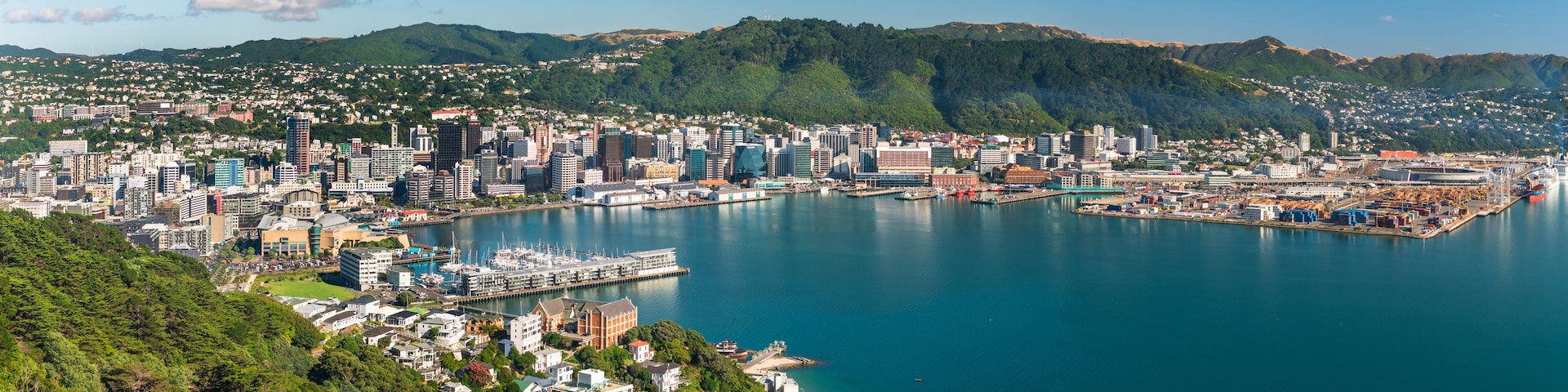 Cidade costeira com um porto de iates, edifícios urbanos densos e colinas ao redor.
