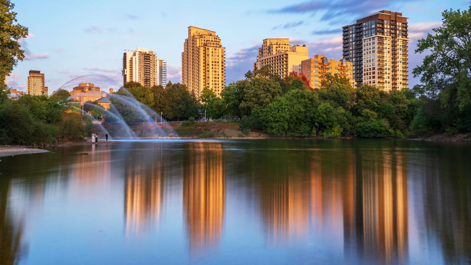 Um lago sereno com uma fonte em primeiro plano, cercado por vegetação exuberante e edifícios altos ao fundo.