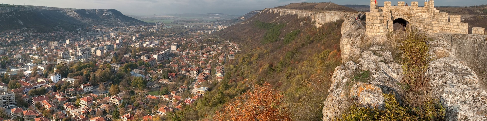 Paisagem montanhosa com um castelo de pedra histórico numa colina, com vista para uma vila.