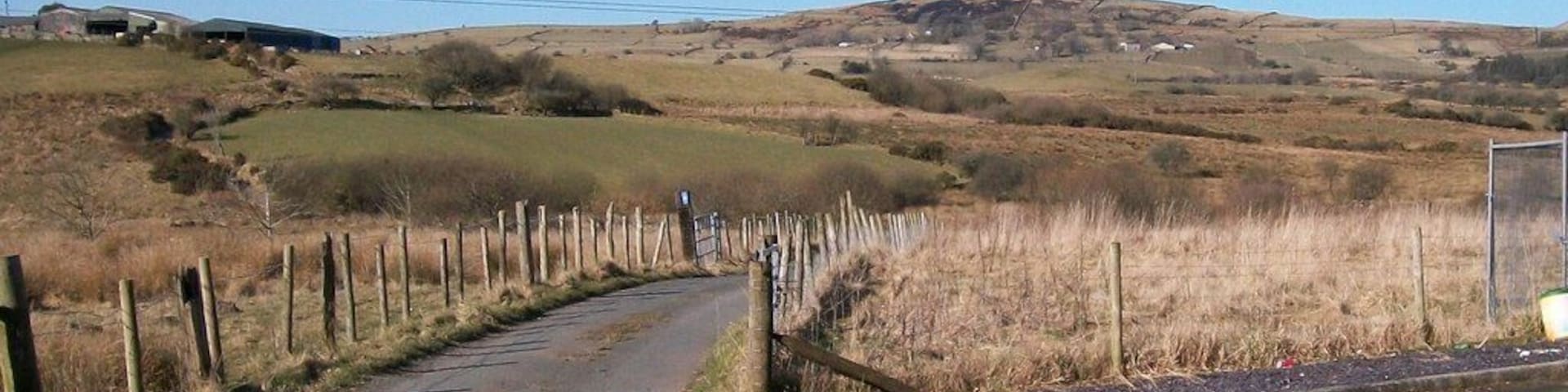 Uma estrada rural serpenteia por uma paisagem montanhosa, com uma cerca de um lado e um céu limpo acima.