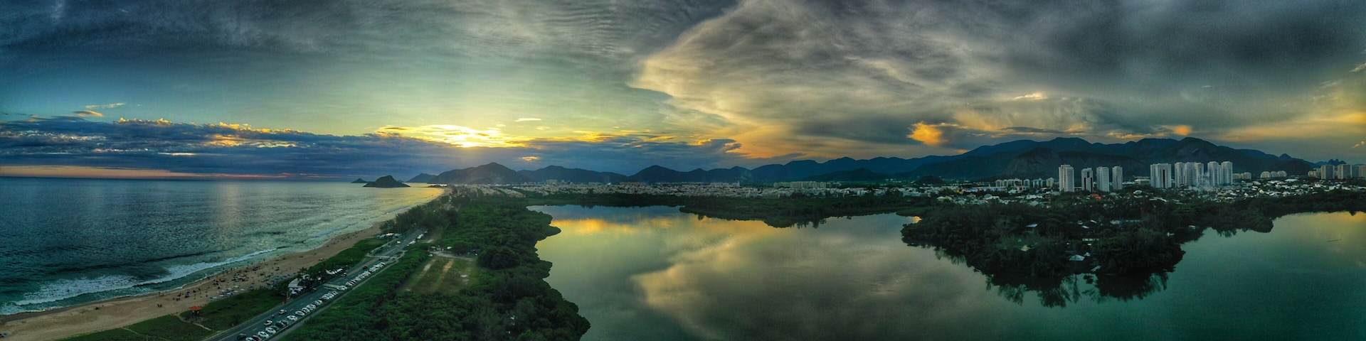 Paisagem costeira com um lago calmo refletindo o céu, uma praia com pessoas e montanhas ao fundo.