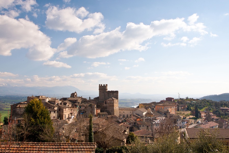 Uma cidade histórica com um castelo imponente, edifícios com telhados de terracota e um céu azul límpido com algumas nuvens.