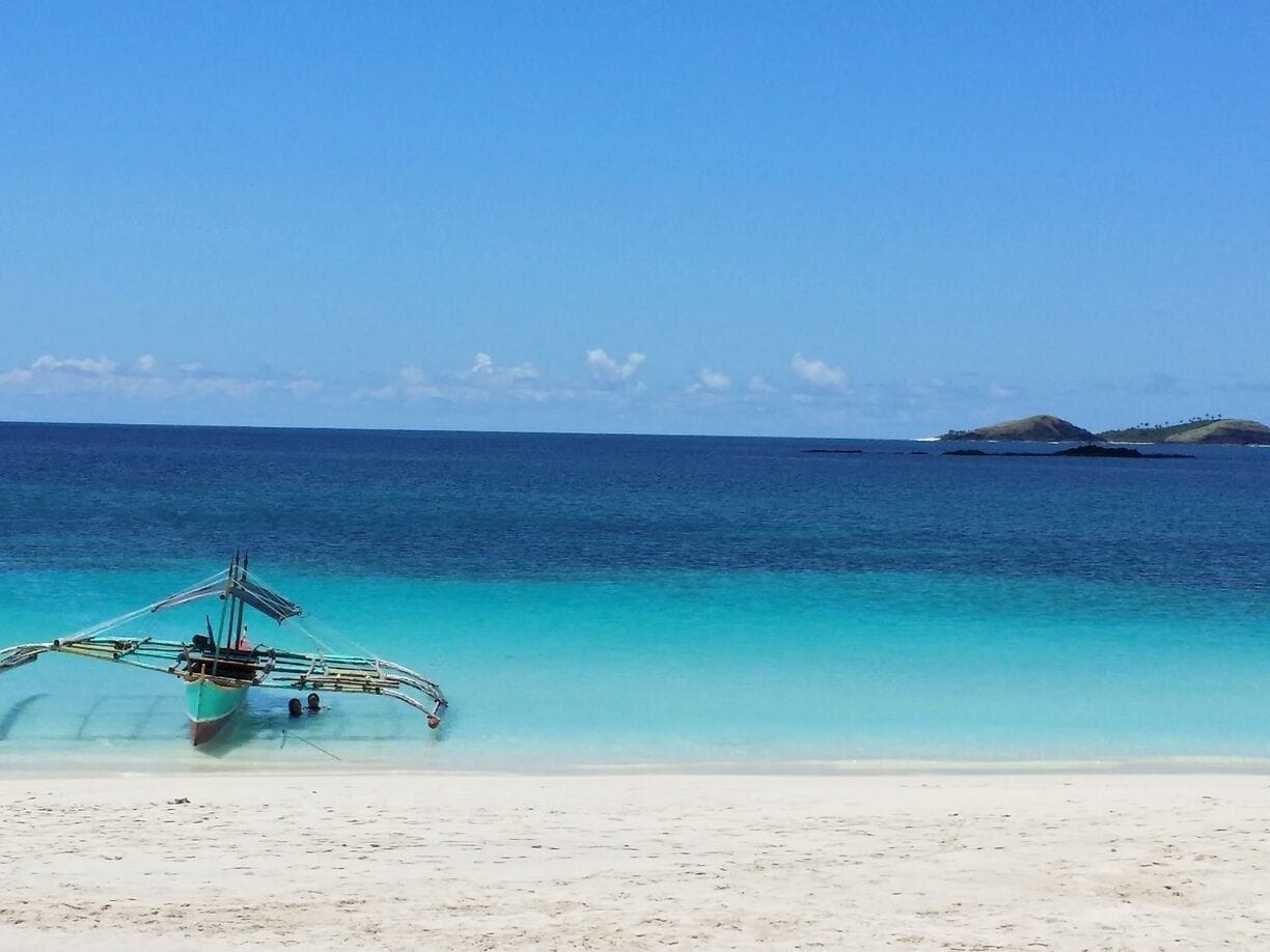 Um barco na praia, com águas turquesa cristalinas e uma ilha ao longe.