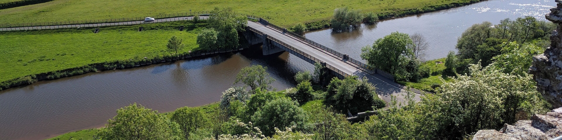 Uma ponte de pedra sobre um rio, rodeada por campos verdes e árvores exuberantes.