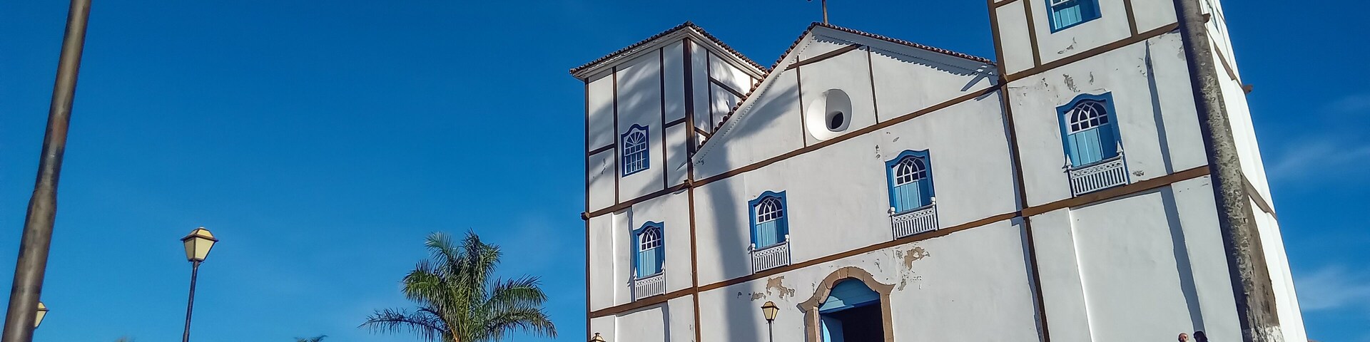 Uma igreja branca com janelas azuis e um campanário, rodeada por palmeiras e um céu azul límpido.