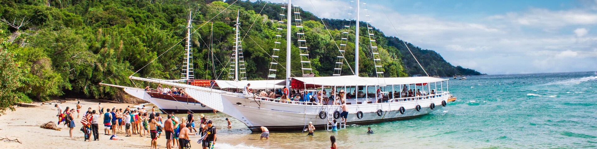 Uma praia com um barco grande atracado perto da costa, cercada por colinas verdes exuberantes e águas azuis cristalinas.