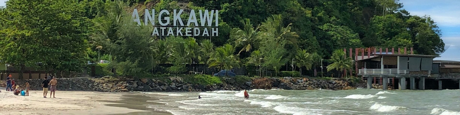 Uma praia com ondas, uma colina arborizada e um edifício com a inscrição 'ANGKAWI ATAKEDAH'.
