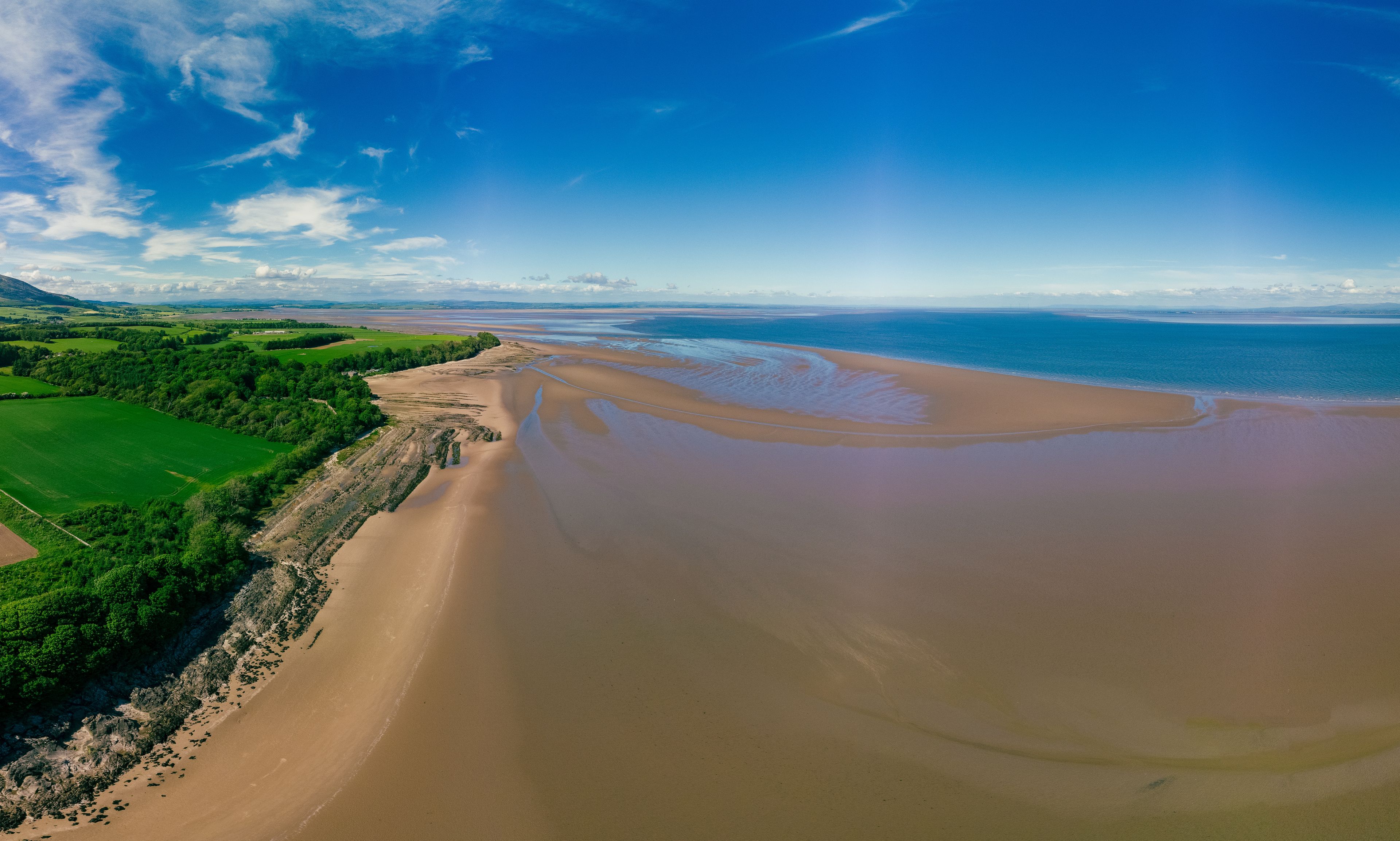 Paisagem costeira com praia de areia, campos verdes e céu azul claro.