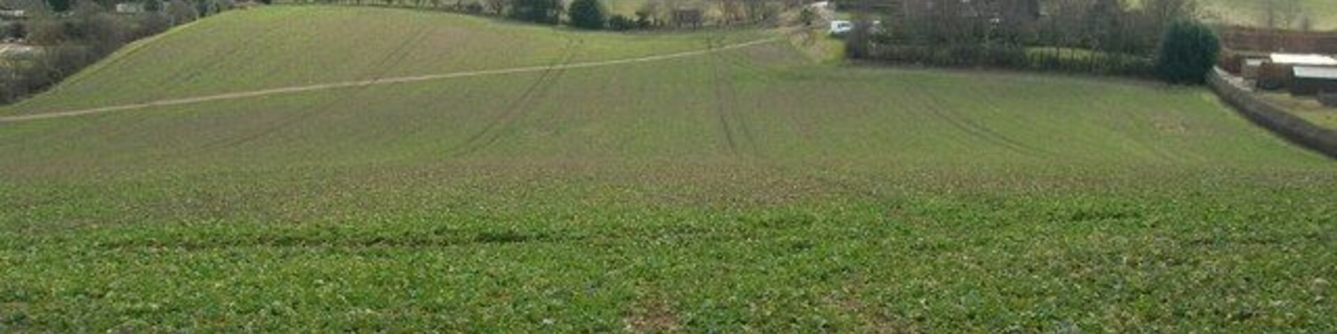 Paisagem rural com um vasto campo aberto, uma aldeia distante e uma colina ao fundo.