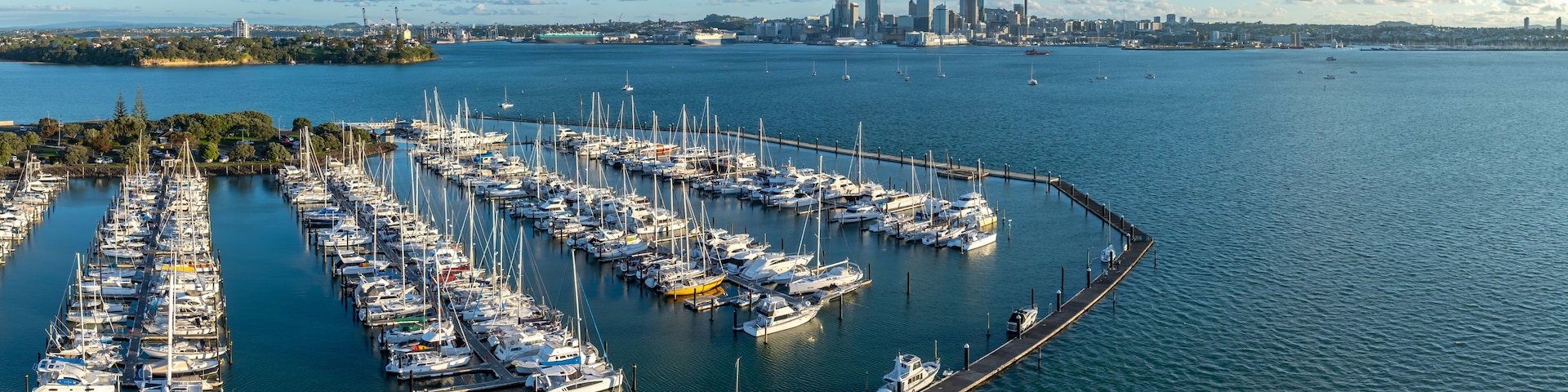 Uma marina com vários barcos atracados numa baía curva, com o skyline de uma cidade ao fundo e céu claro.