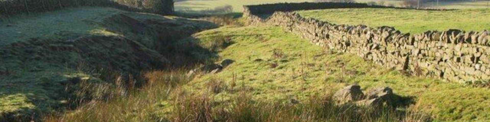Paisagem rural com muros de pedra, campos gramados e edifícios ao longe.