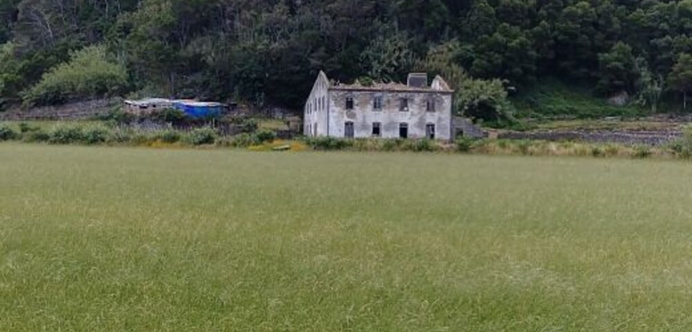 Paisagem rural com um grande campo em primeiro plano e uma casa numa colina ao fundo.