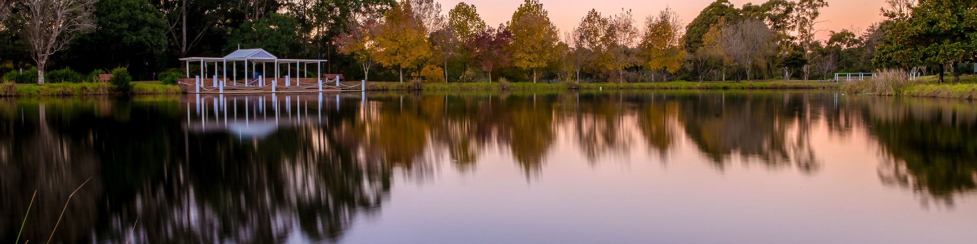 Um lago calmo com um coreto, rodeado por árvores com folhagem outonal, refletindo o céu.