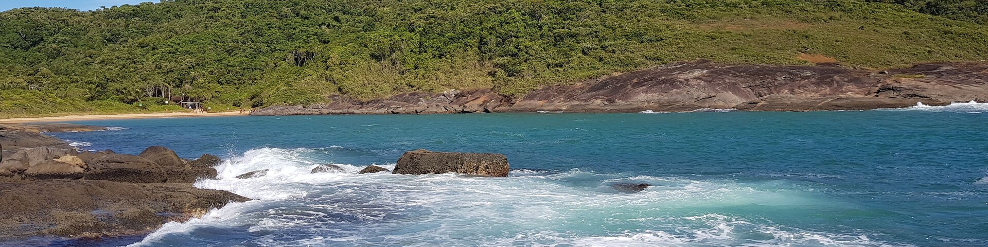 Paisagem costeira com costas rochosas, águas azul-claras e uma colina arborizada ao fundo.