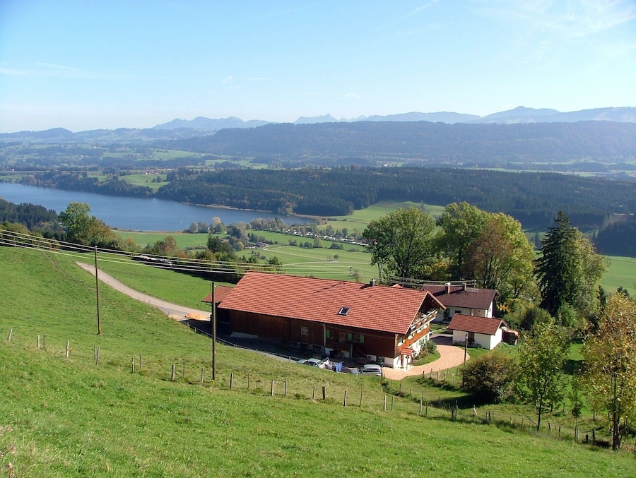Paisagem rural com um lago, colinas onduladas e uma casa de telhado vermelho.
