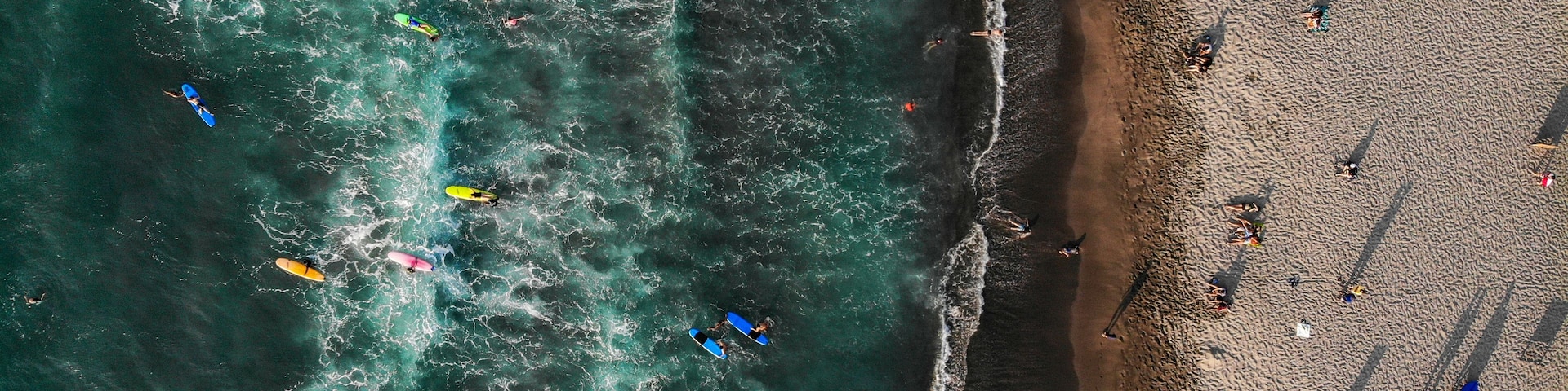 Vista aérea de uma praia com surfistas e nadadores na água, e pessoas caminhando na areia.