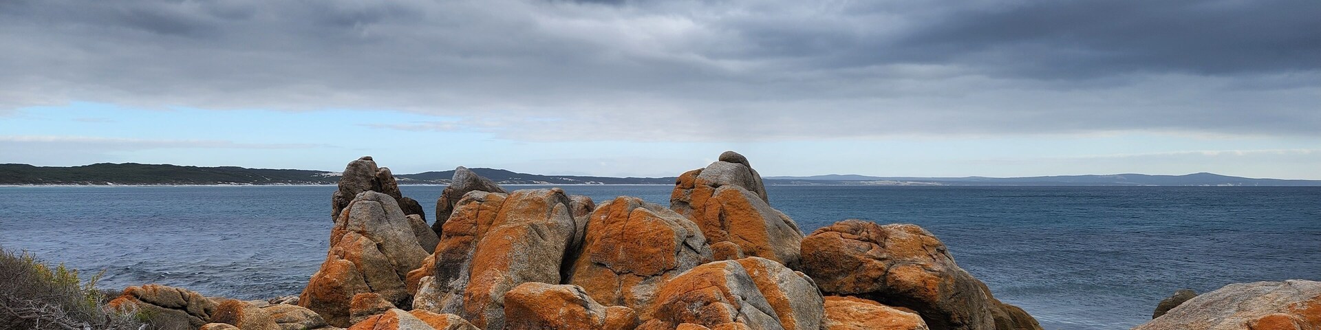 Litoral rochoso com grandes pedras cobertas de líquen alaranjado e vista para o mar sob um céu nublado.