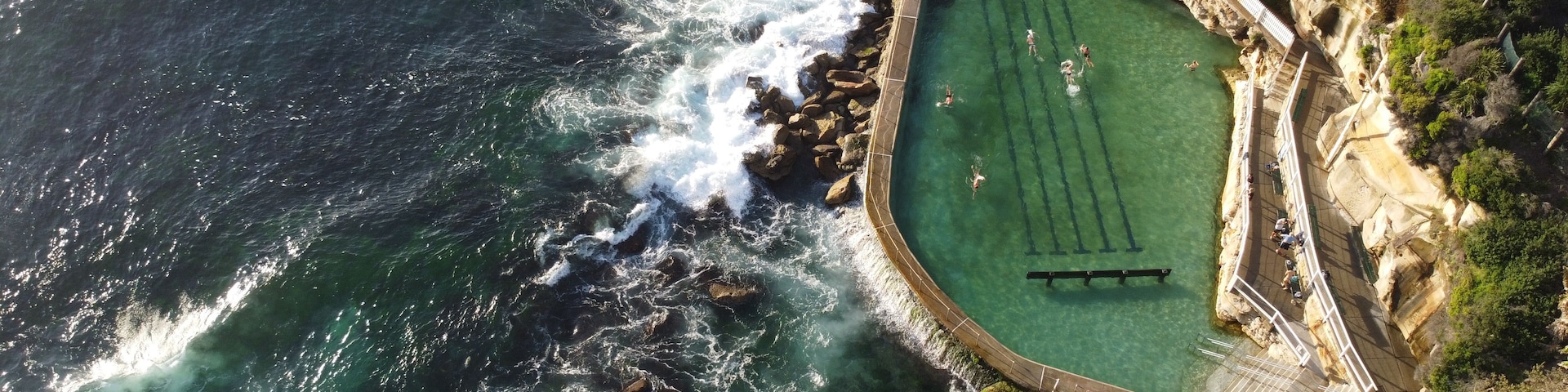 Vista aérea de uma área costeira com uma piscina límpida, cercada por falésias rochosas e ondas quebrando na costa.