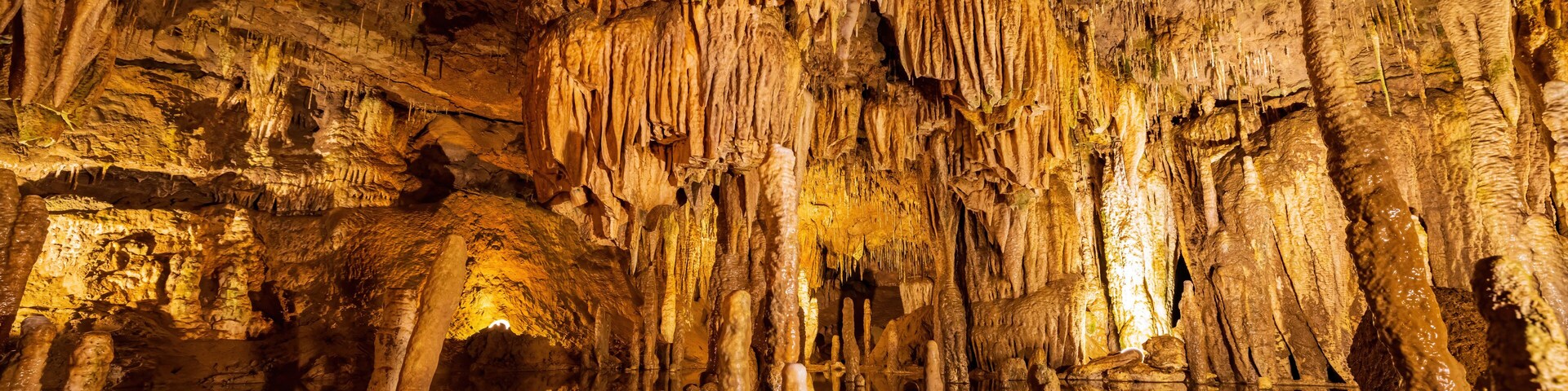 Caverna com estalactites e estalagmites, reflexos de água e uma abertura.