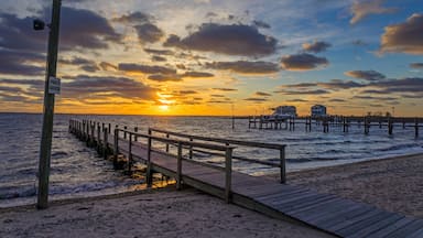 Um píer de madeira que se estende para o oceano ao pôr do sol, com uma placa indicando 'Public Access', uma praia e uma casa ao longe.