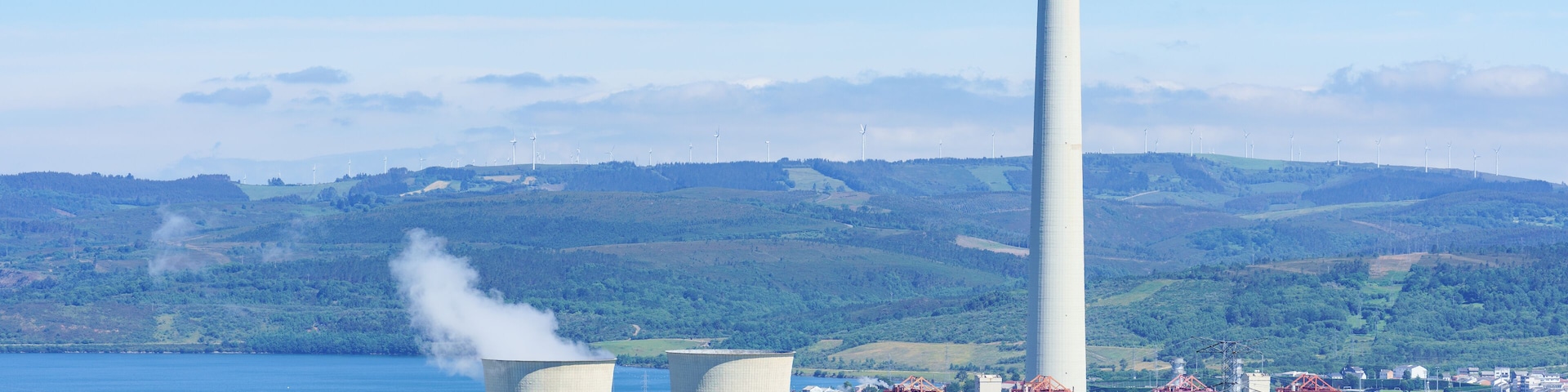 Uma central elétrica com torres de resfriamento e uma chaminé alta, rodeada por colinas e um corpo d'água.