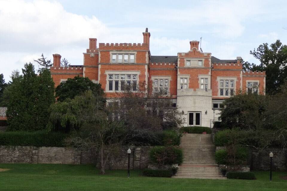 Um edifício grande e histórico de tijolos, com fachada simétrica, entrada central e um gramado bem cuidado, com muro de pedra e degraus que levam à entrada.