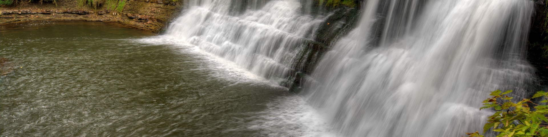 Uma cascata em vários níveis deságua em um rio calmo, cercado por vegetação exuberante e estruturas de pedra.