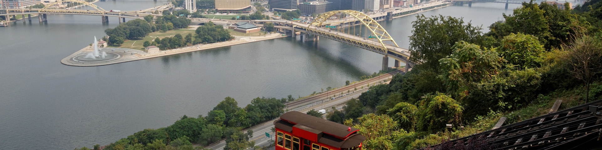 Um teleférico vermelho sobre trilhos, um rio, horizonte urbano com edifícios altos e uma ponte.