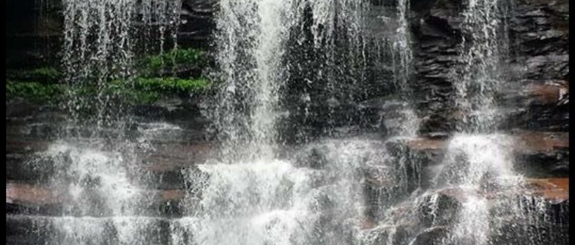 Uma cascata despencando sobre rochas em área arborizada, com uma pessoa próxima à base.