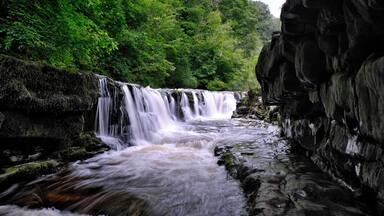 Uma cascata despencando sobre terreno rochoso, cercada por vegetação verde densa.
