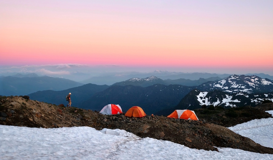 Um caminhante numa trilha de montanha com tendas e neve.
