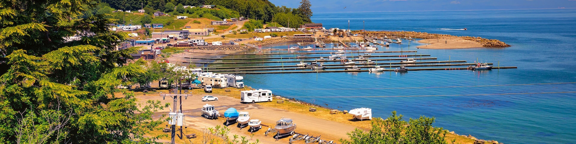 Uma área costeira com um porto de iates, barcos, veículos recreativos e uma encosta arborizada.
