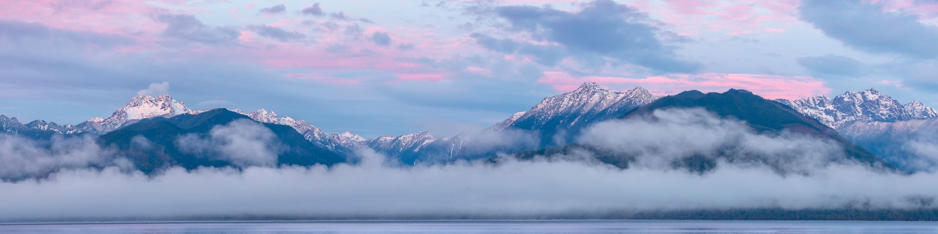 Uma cordilheira com picos nevados, um lago em primeiro plano e um céu com tons de rosa e azul.